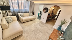 Sala de estar com sofá bege, mesa de centro e televisão. É possível ver, através de um arco, uma área de jantar com mesa e cadeiras.