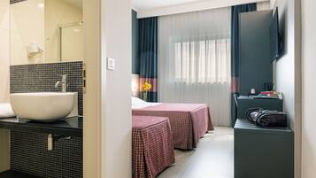 Quarto de hotel com uma pia grande no banheiro, um espelho, uma cama com colcha xadrez, uma mesinha de cabeceira, uma televisão na parede e uma mala em um banco.
