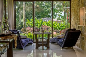 Quarto com mesa de vidro, dois sofás azuis, parede de pedra e vista para vegetação exuberante.