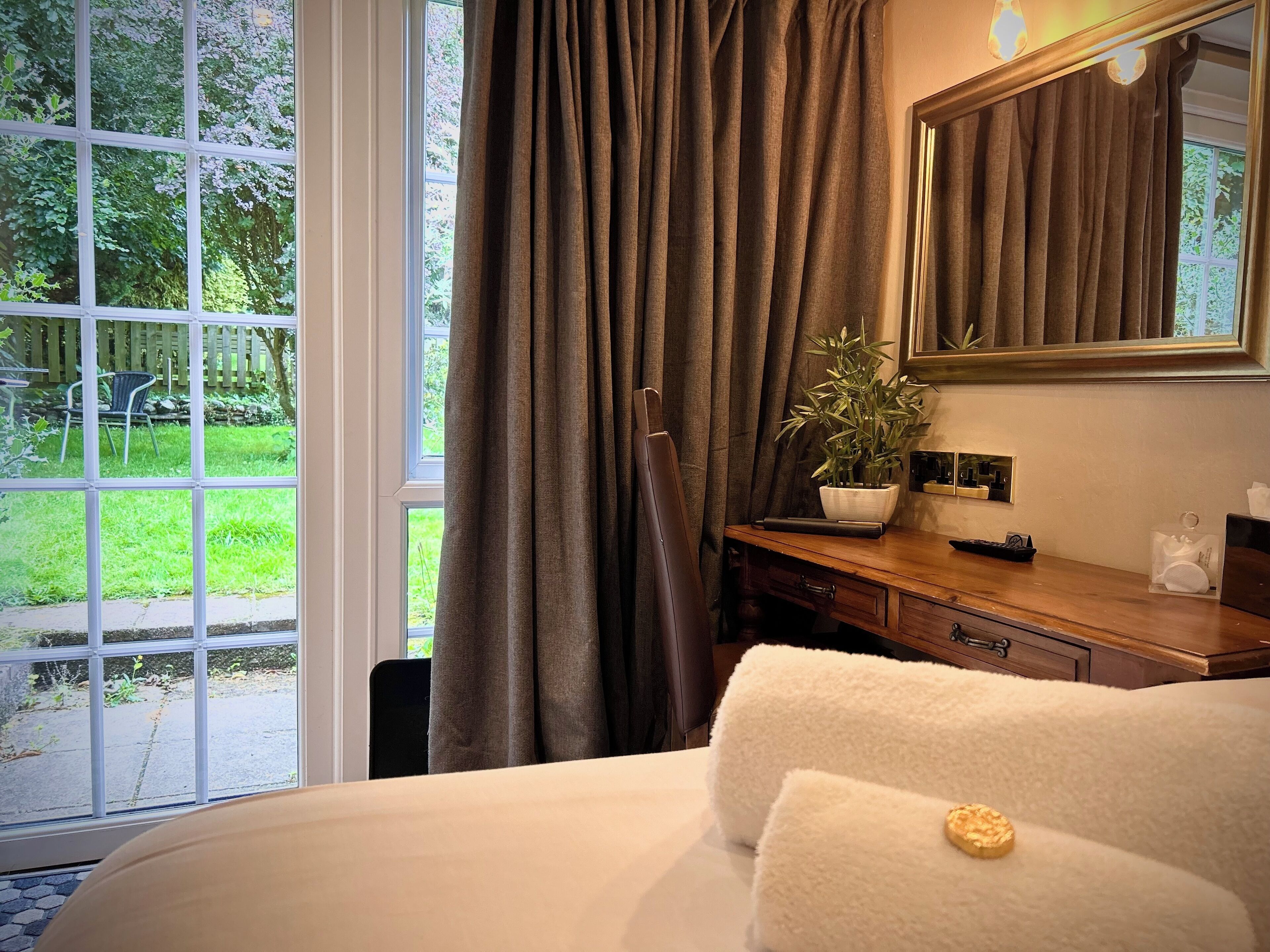 Quarto de hotel com uma janela grande, uma escrivaninha de madeira, um espelho, uma planta e uma cama com roupa de cama branca.