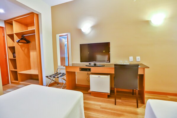 Quarto de hotel com escrivaninha de madeira, televisão, mini-frigorífico, cadeira, cama e guarda-roupa.