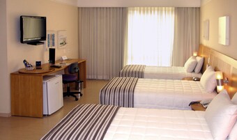 Quarto de hotel com duas camas, uma escrivaninha, televisão e janela com cortinas.