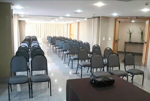 Uma sala de conferências com fileiras de cadeiras, uma mesa comprida e uma porta com uma placa.