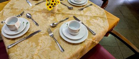 Mesa posta para quatro com pratos, copos e talheres brancos, um vaso com flores amarelas e toalha de mesa amarela.