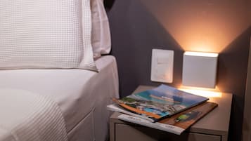 Cama bem arrumada com edredom e manta brancos. Uma revista está sobre a mesa de cabeceira, ao lado do interruptor de luz fixado na parede.