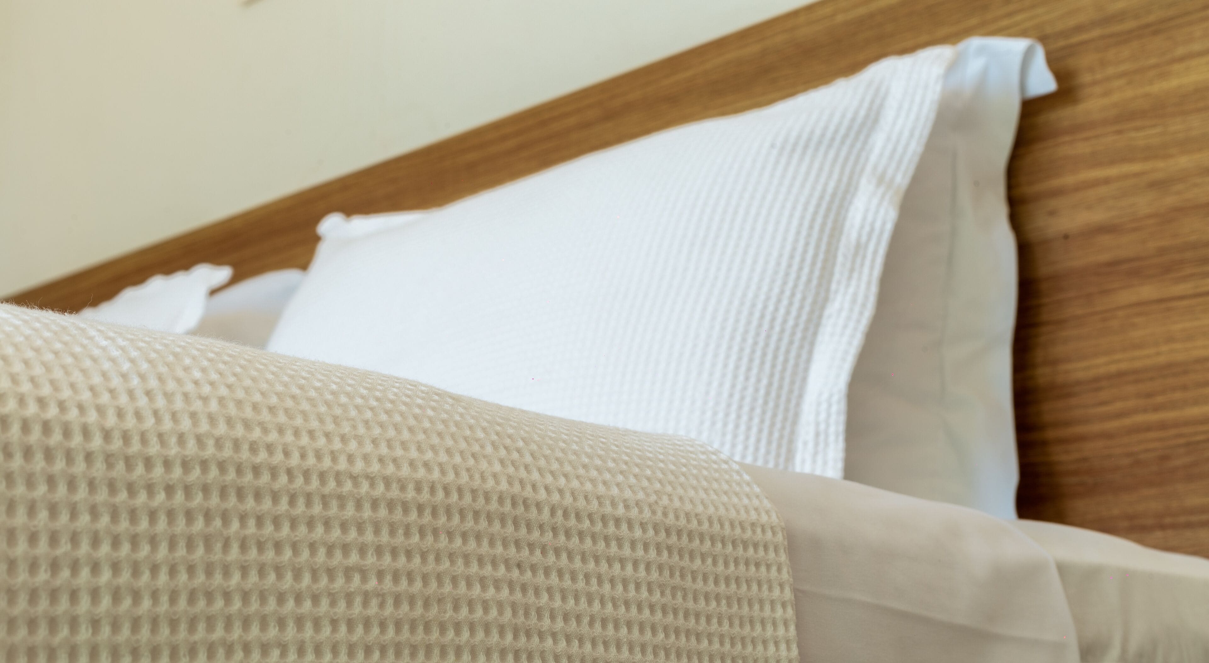 Uma cama com cabeceira de madeira, almofadas brancas e um cobertor bege.