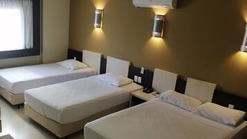 Quarto de hotel com três camas de solteiro, cada uma com lençóis brancos e cabeceiras. Há luminárias fixadas na parede e um aparelho de ar condicionado.