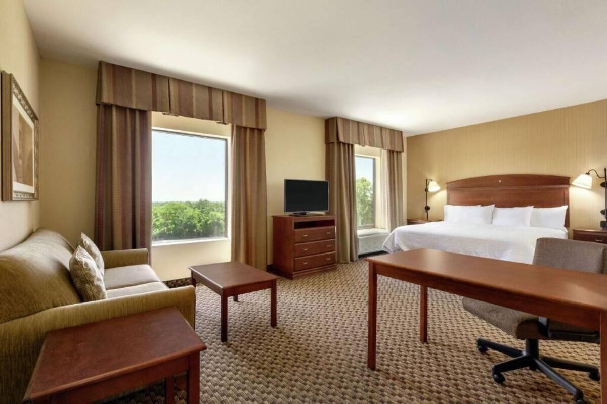 Quarto de hotel com cama, televisão, escrivaninha, poltrona, sofá e janelas amplas.