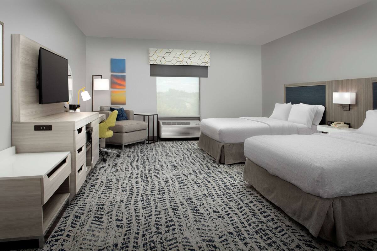 Quarto de hotel com uma escrivaninha, duas camas, uma televisão, uma janela e um tapete estampado.