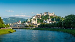 Um castelo histórico numa colina com vista para um rio, rodeado por vegetação exuberante e uma pequena ponte.