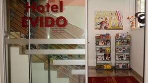 Entrada de hotel com escadaria, balcão de recepção e uma parede com um cartaz e uma estante de livros.