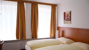 Quarto de hotel com cama, duas almofadas, radiador e janela com cortinas.
