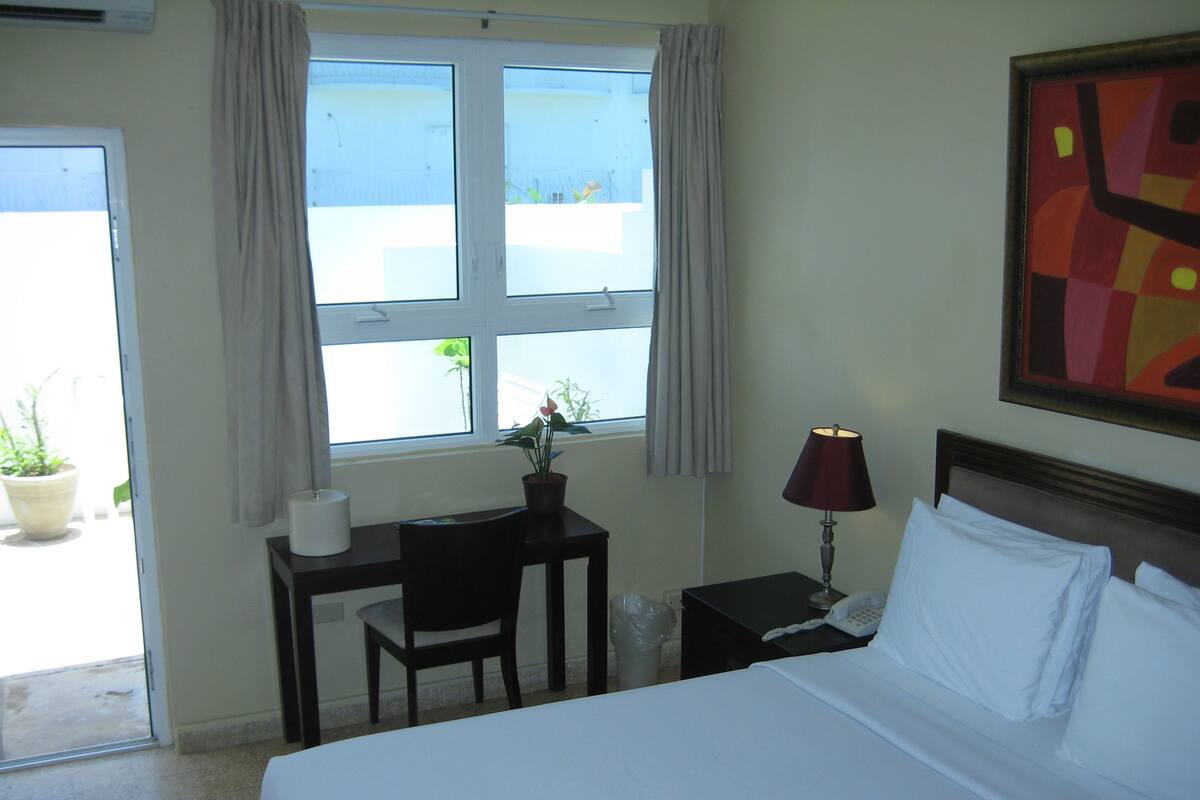 Quarto de hotel com cama, escrivaninha, cadeira, luminária, telefone e janela com cortinas.