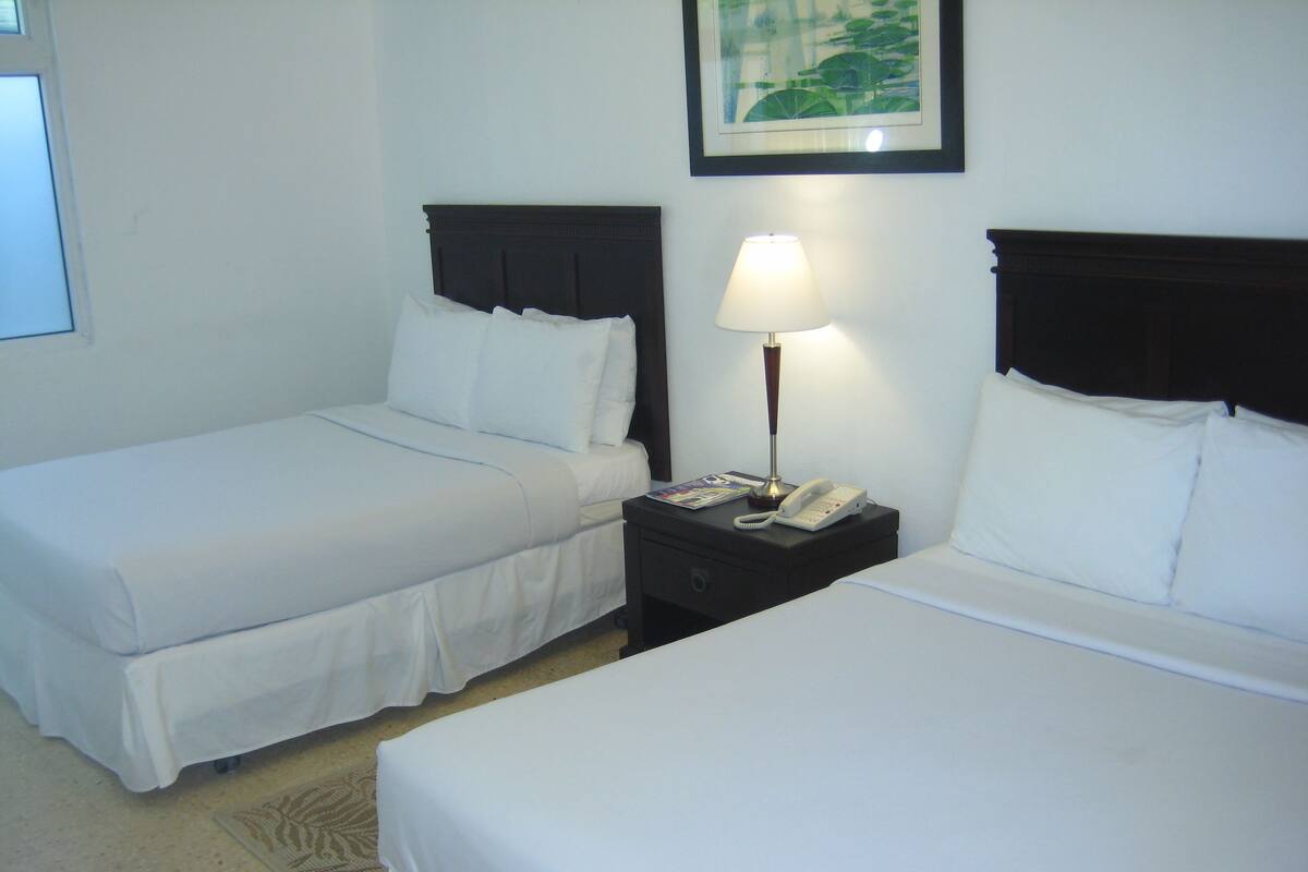 Quarto de hotel com duas camas de solteiro, mesa de cabeceira com abajur e telefone, e um quadro na parede.