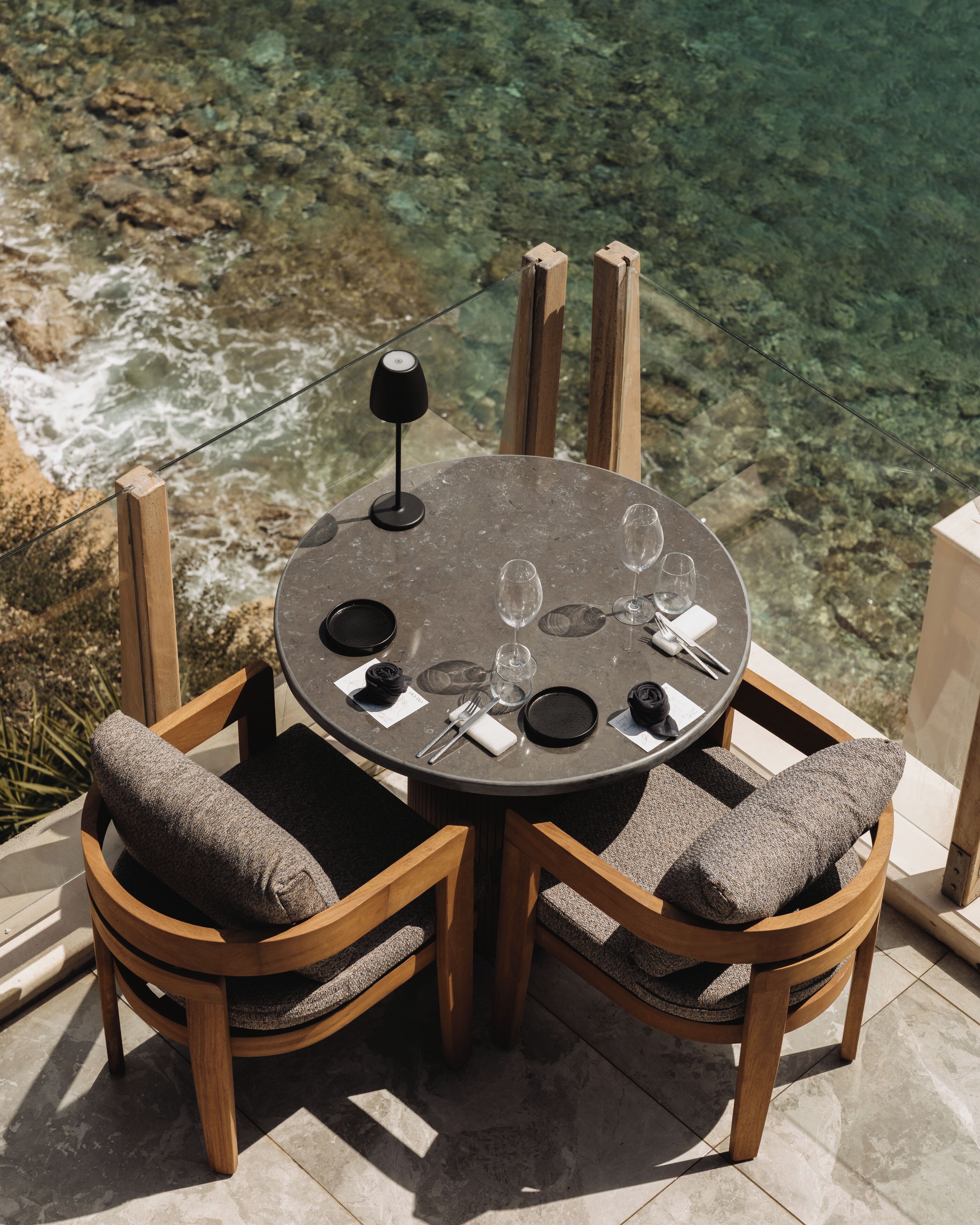 Uma mesa redonda com quatro cadeiras, uma taça de vinho e uma vela em um terraço com vista para a costa rochosa.
