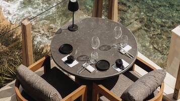 Uma mesa redonda com quatro cadeiras, uma taça de vinho e uma vela em um terraço com vista para a costa rochosa.