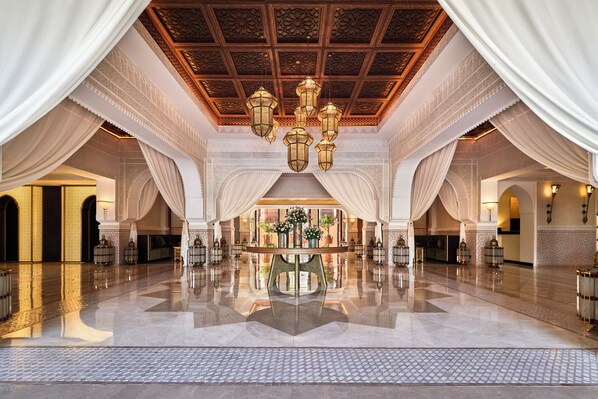 Um hall amplo com teto alto, lustres ornamentados e uma mesa central com arranjos florais.