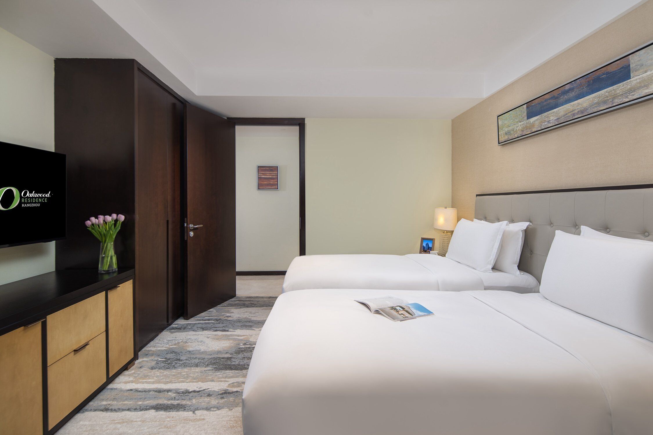 Quarto de hotel com duas camas, televisão, secretária com um vaso de flores e um quadro na parede.