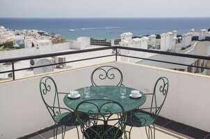 Uma varanda com mesa e cadeiras, com vista para uma paisagem urbana costeira e o mar ao fundo.