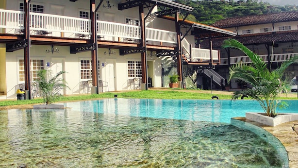 Uma piscina translúcida com borda de pedra, um edifício branco com varandas e uma escadaria que leva aos andares superiores.