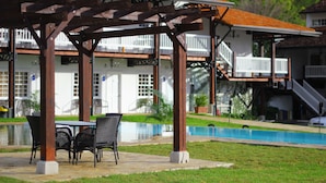 Uma área de estar coberta ao ar livre com piscina e um edifício ao fundo.