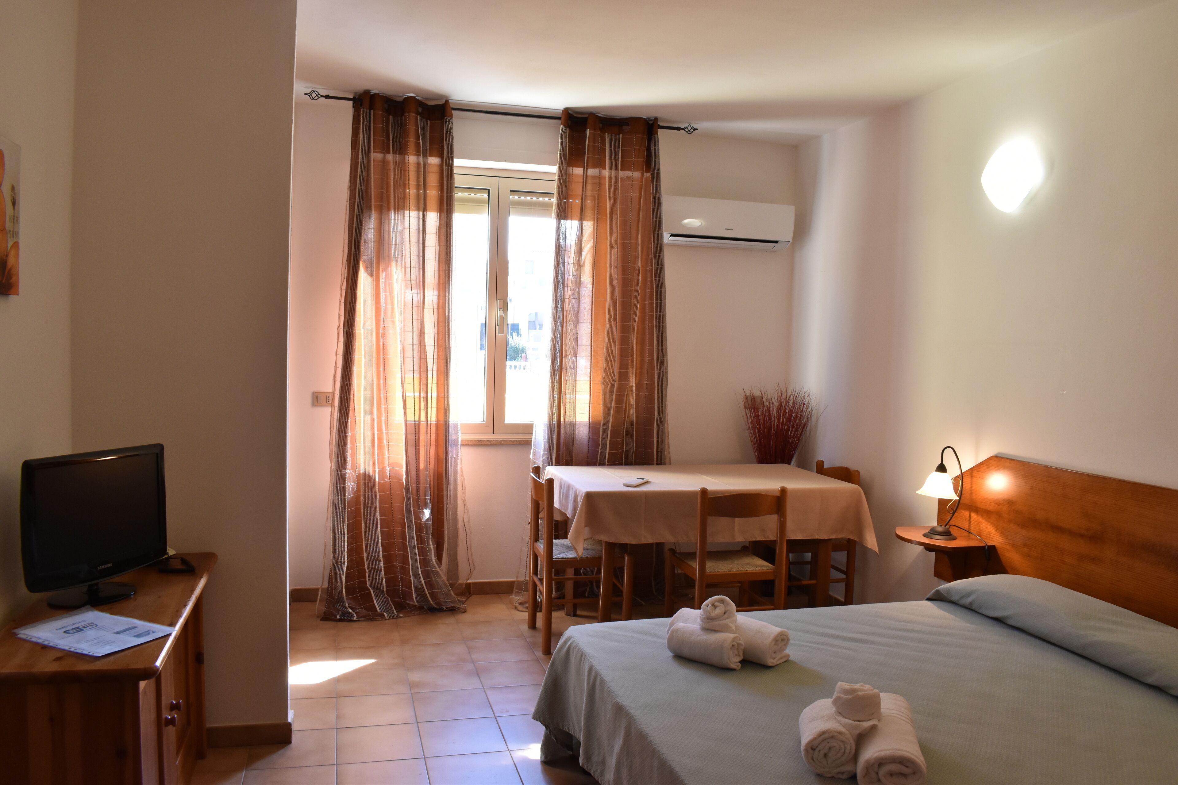 Quarto de hotel com cama, escrivaninha com computador, mesa com cadeiras e janela com cortinas.
