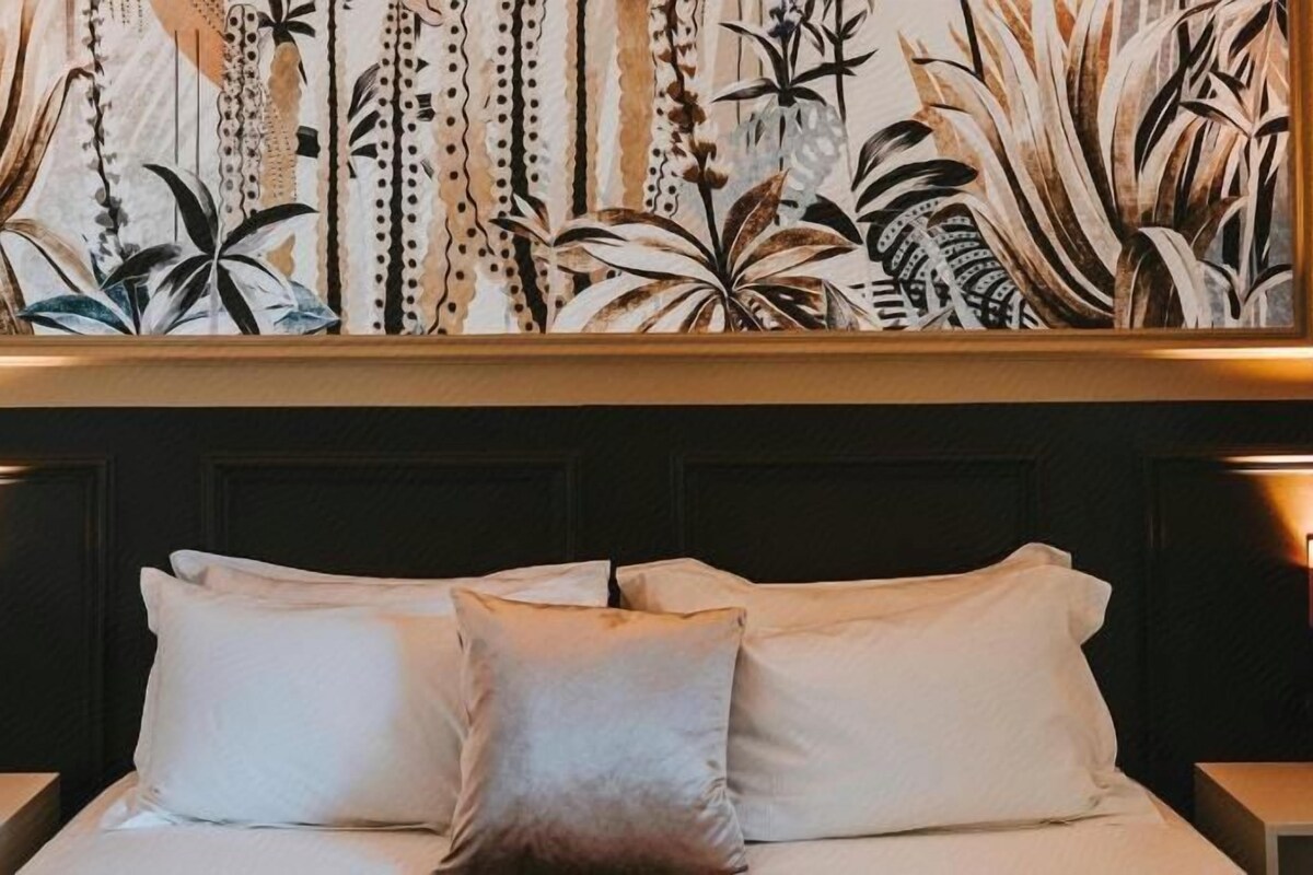 Cama com lençóis brancos e um único travesseiro decorativo, com duas mesas de cabeceira e luminárias fixadas na parede.
