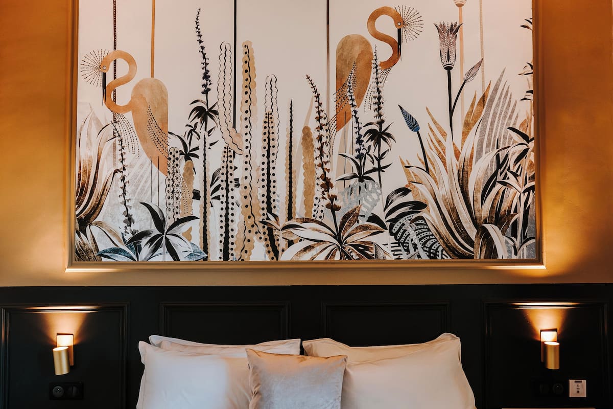 Cama com lençóis brancos, duas mesas de cabeceira e uma obra de arte na parede retratando pássaros e plantas estilizados.