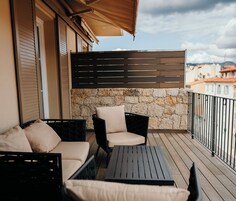 Uma varanda com mobiliário externo, uma parede de pedra e vista para edifícios ao fundo.