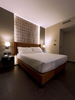 Quarto de hotel com uma cama grande, duas mesas de cabeceira, um criado-mudo, uma luminária na parede e uma janela com persianas.