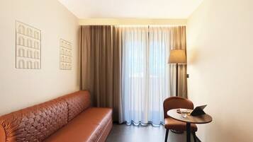 Quarto de hotel com um sofá de couro marrom, uma mesa redonda pequena com um abajur e uma janela com cortinas.