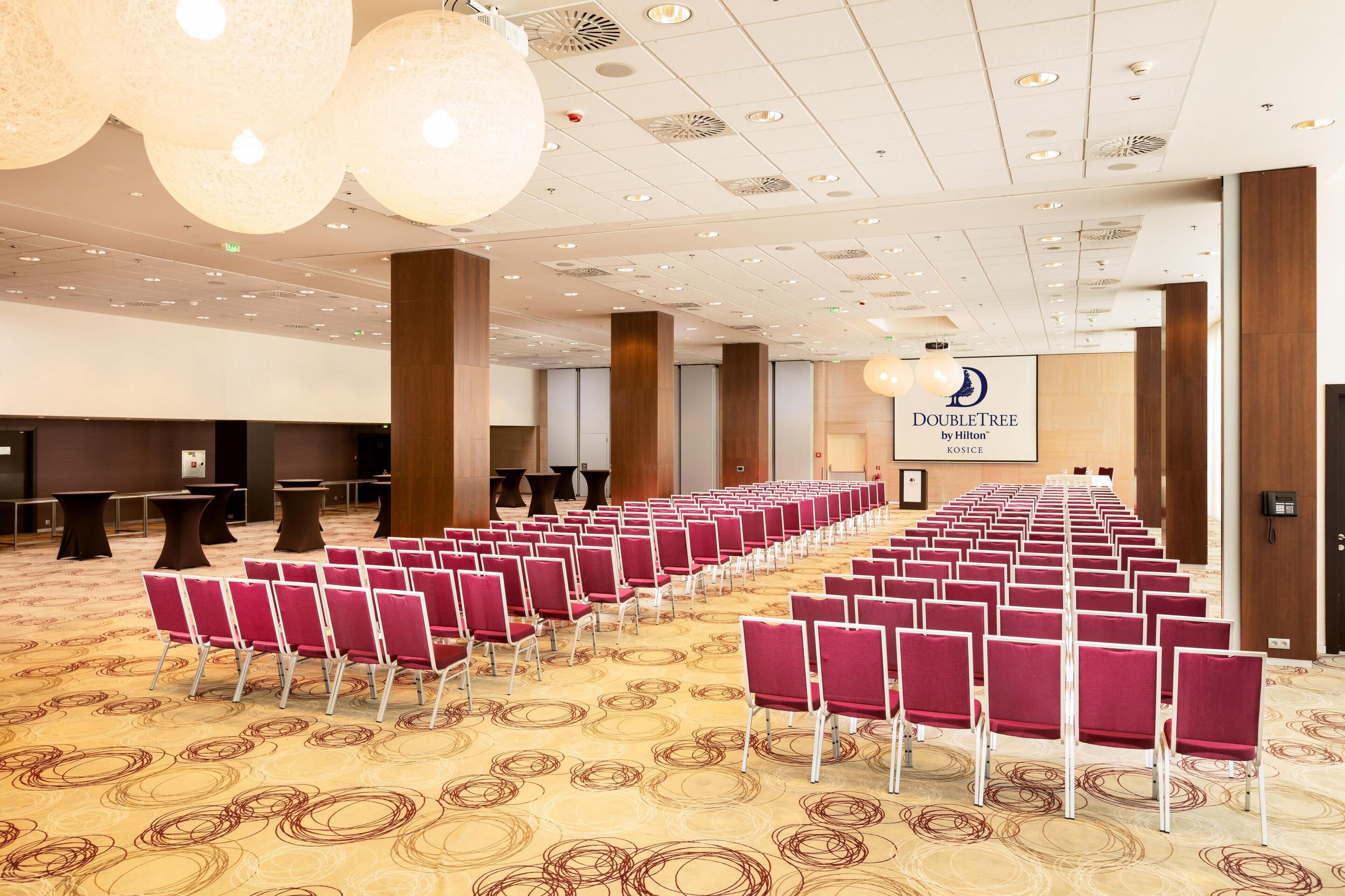Uma sala de conferências com fileiras de cadeiras vermelhas, piso coberto por carpete e uma tela grande exibindo o logotipo da DoubleTree by Hilton.