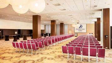 Uma sala de conferências com fileiras de cadeiras vermelhas, piso coberto por carpete e uma tela grande exibindo o logotipo da DoubleTree by Hilton.