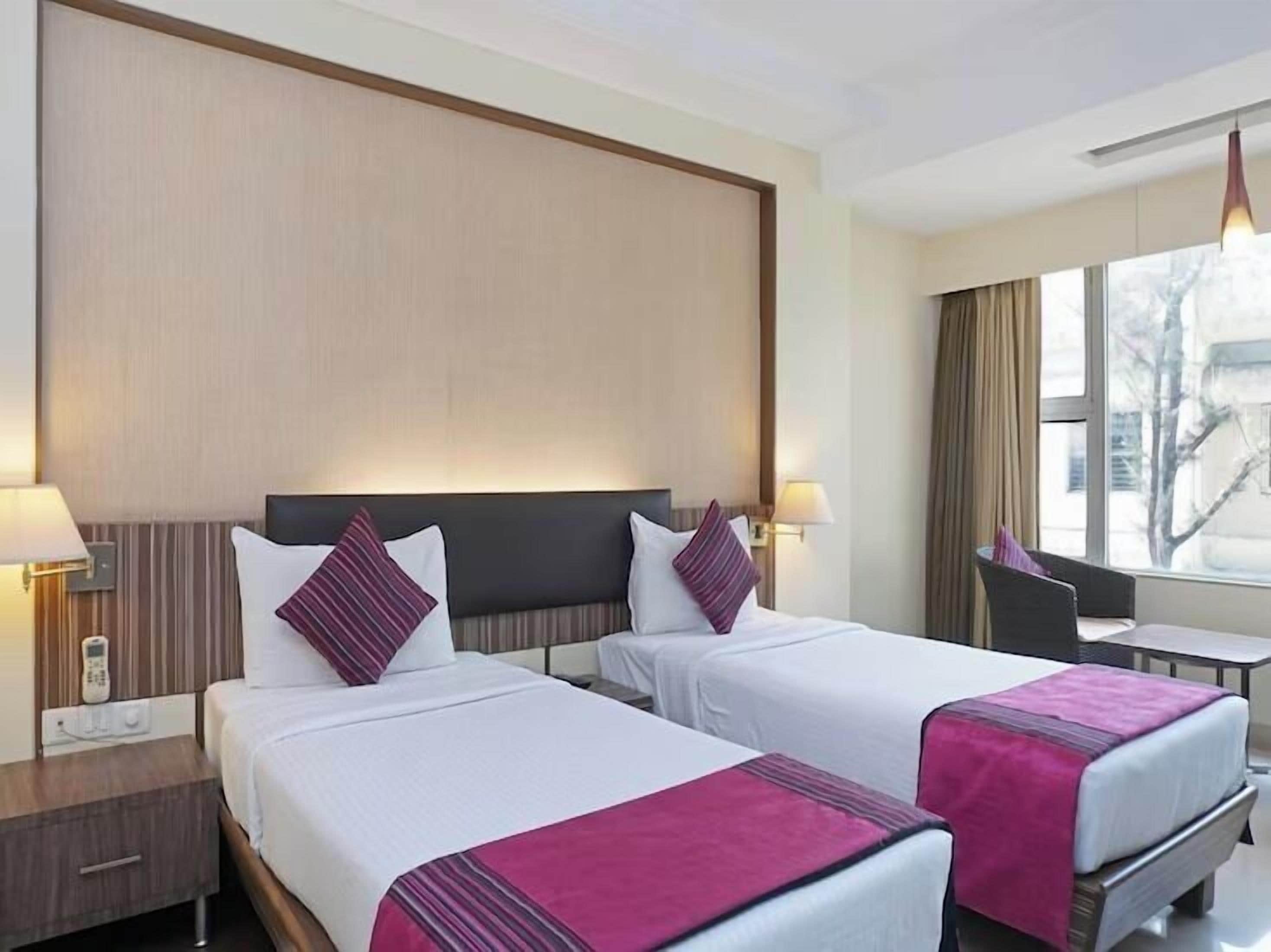 Quarto de hotel com duas camas, mesa de cabeceira, luminária, janela com cortinas e vista para o exterior.