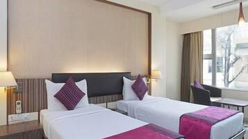 Quarto de hotel com duas camas, mesa de cabeceira, luminária, janela com cortinas e vista para o exterior.