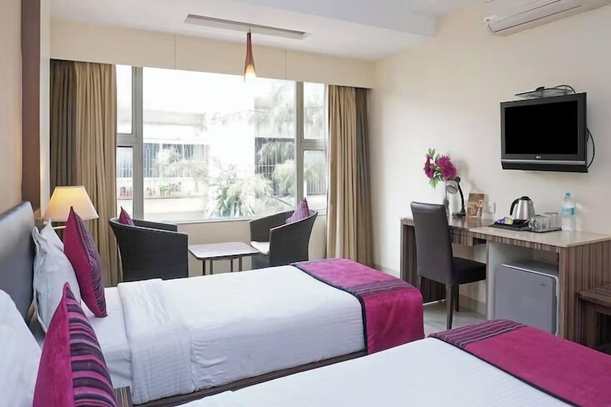 Quarto de hotel com duas camas, uma escrivaninha, uma cadeira, uma televisão e uma janela com cortinas.