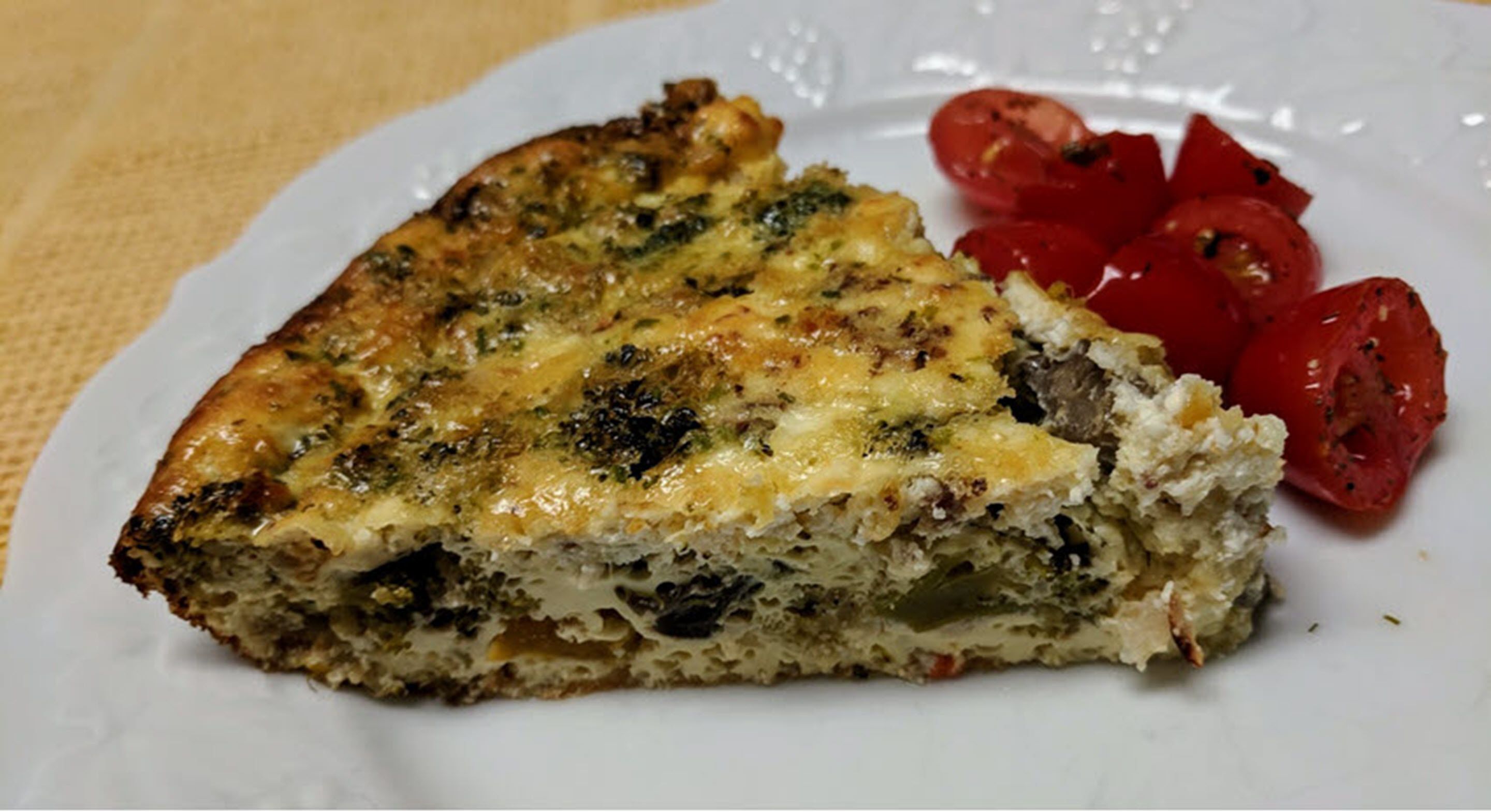 Uma fatia de quiche com crosta dourada e recheio verde, acompanhada de tomate assado em um prato branco.