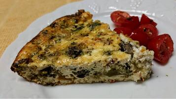 Uma fatia de quiche com crosta dourada e recheio verde, acompanhada de tomate assado em um prato branco.