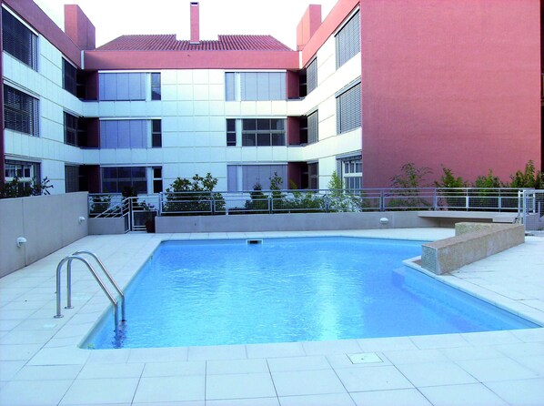 Uma piscina com um deck revestido, cercada por um edifício com várias andares, varandas e uma parede vermelha.