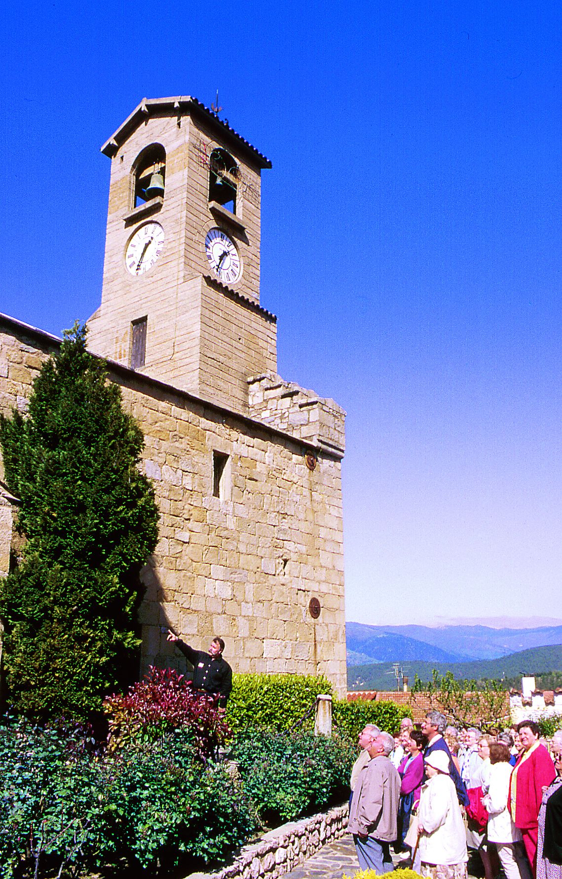 Um edifício histórico de pedra com uma torre do relógio, cercado por vegetação exuberante e um céu azul límpido.