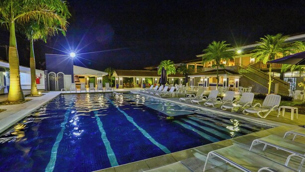 Área de piscina à noite, com espreguiçadeiras e palmeiras.