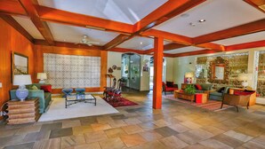 Ampla sala de estar com pé direito alto, vigas de madeira e parede estampada. Há vários espaços de estar com sofás e poltronas, uma mesa de centro e uma lareira.