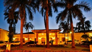 Um hotel com entrada bem iluminada, palmeiras e carros estacionados.