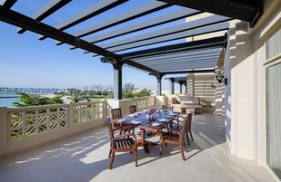 Uma área de refeições ao ar livre coberta, com uma mesa posta para quatro pessoas, cadeiras e vista para o horizonte da cidade.