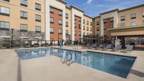 Piscina com espreguiçadeiras e guarda-sóis, rodeada por edifícios altos com varandas.