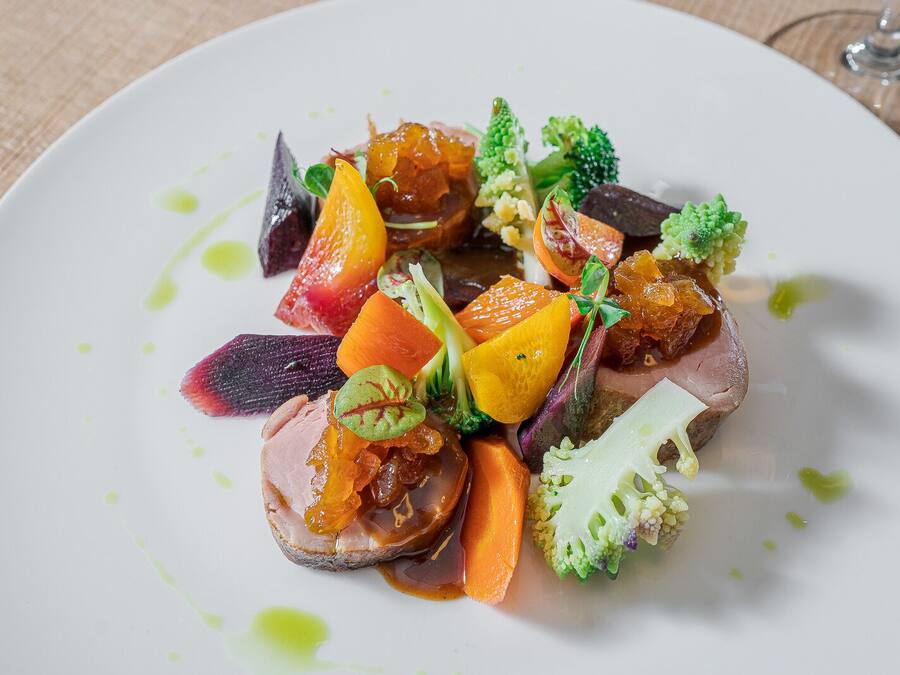 Um prato branco com uma variedade de legumes coloridos e um pedaço de carne, possivelmente um bife, decorado com folhas verdes.