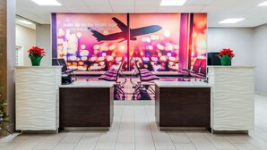 Uma área de recepção com um grande mural com tema de aeroporto, dois balcões de atendimento e flores vermelhas decorativas.