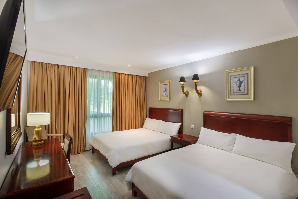 Quarto de hotel com duas camas, uma escrivaninha com abajur, um espelho e duas luminárias de parede.