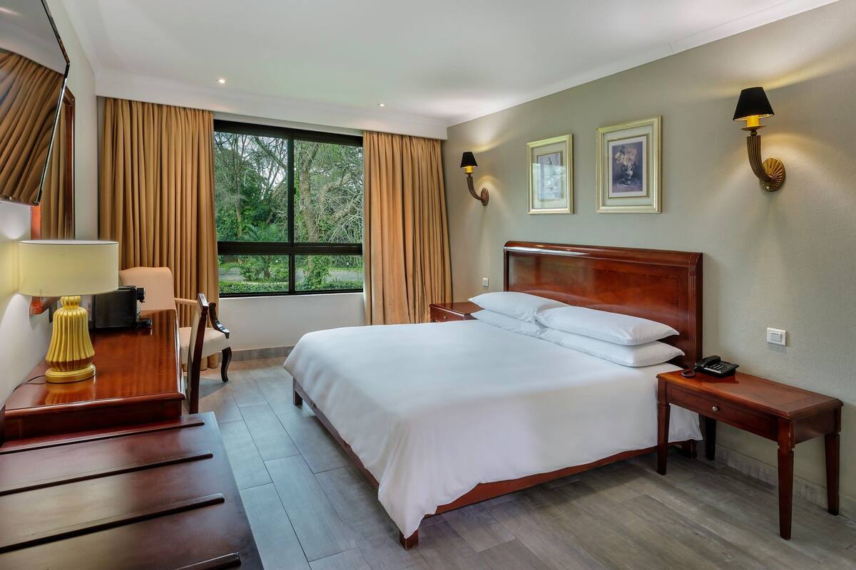 Quarto de hotel com uma cama grande, uma mesa com luminária, uma cadeira e vista para o verde lá fora.