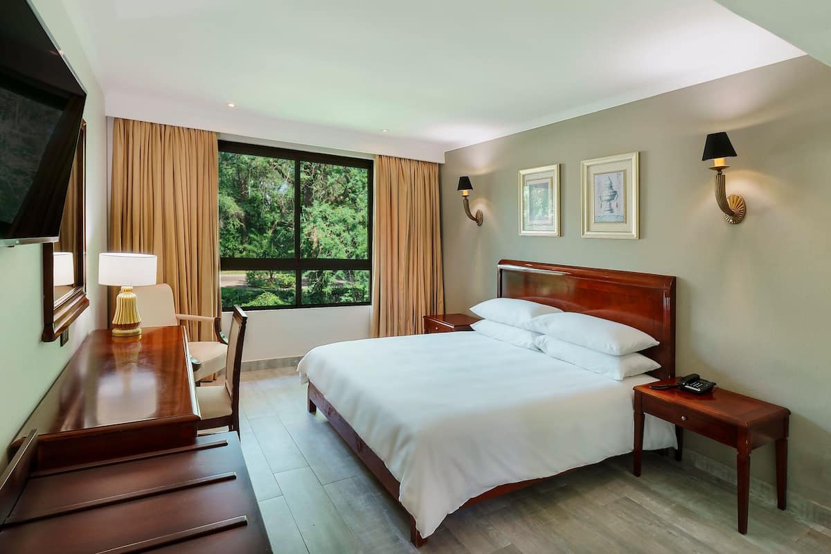 Quarto de hotel com cama, mesa de cabeceira, abajur e televisão sobre suporte.
